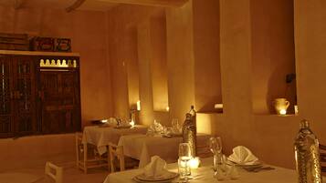 Uma sala de jantar com pouca luz, com mesas preparadas para dois, com velas, copos de vinho e uma garrafa decorativa.