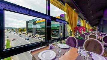 Restaurante com vista para uma rua movimentada e edifícios modernos. Mesas dispostas com pratos e talheres brancos.
