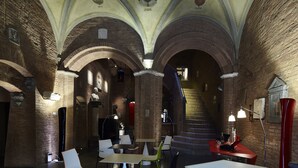 Um edifício histórico com tetos abobadados, paredes de tijolo e uma escadaria que leva a um andar superior. Mesas e cadeiras estão dispostas na área inferior, e há escudos decorativos no teto.
