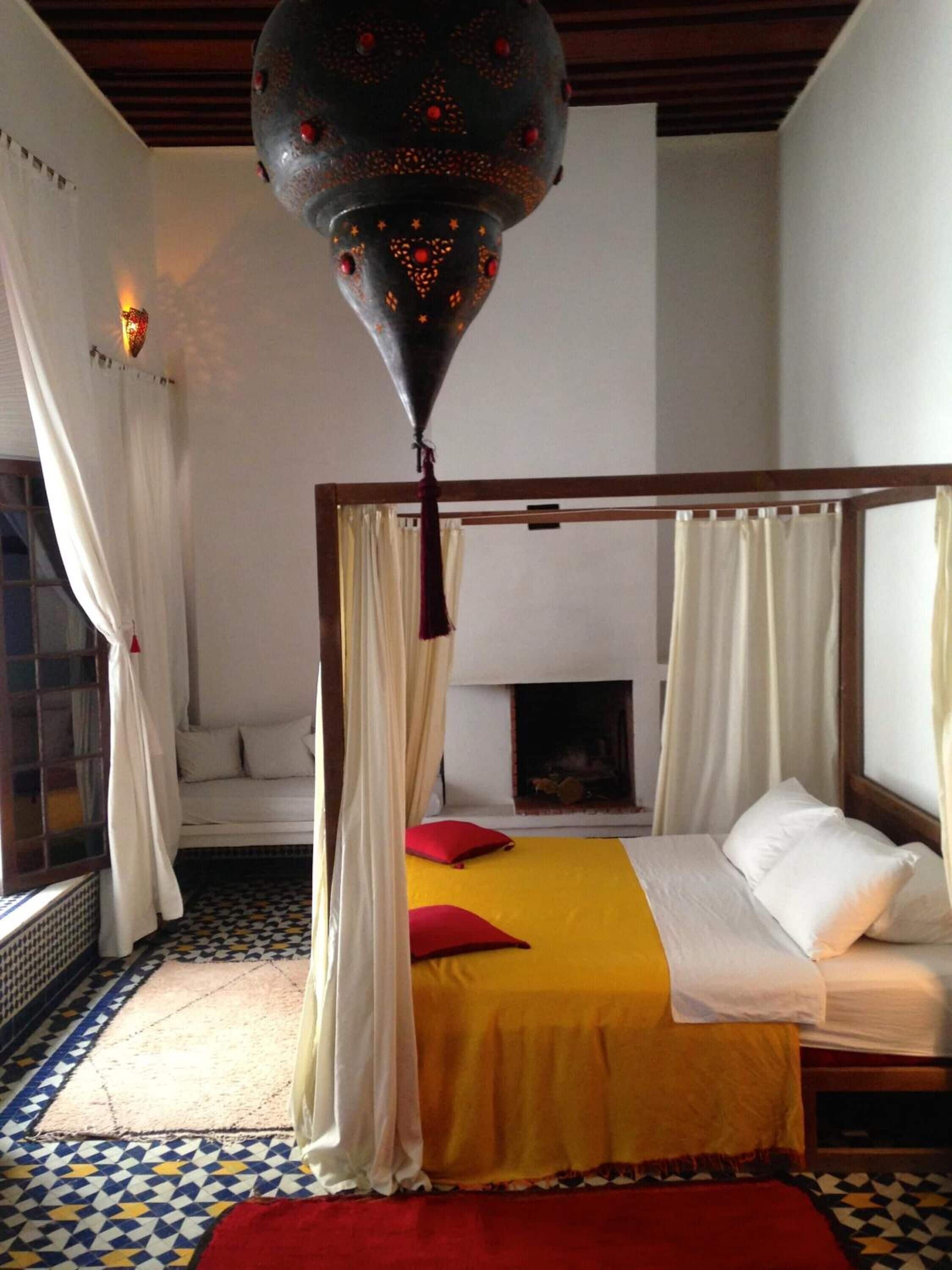 Uma cama com dossel, coberta por um edredom amarelo e travesseiros vermelhos, uma lareira e um chão com padrão.