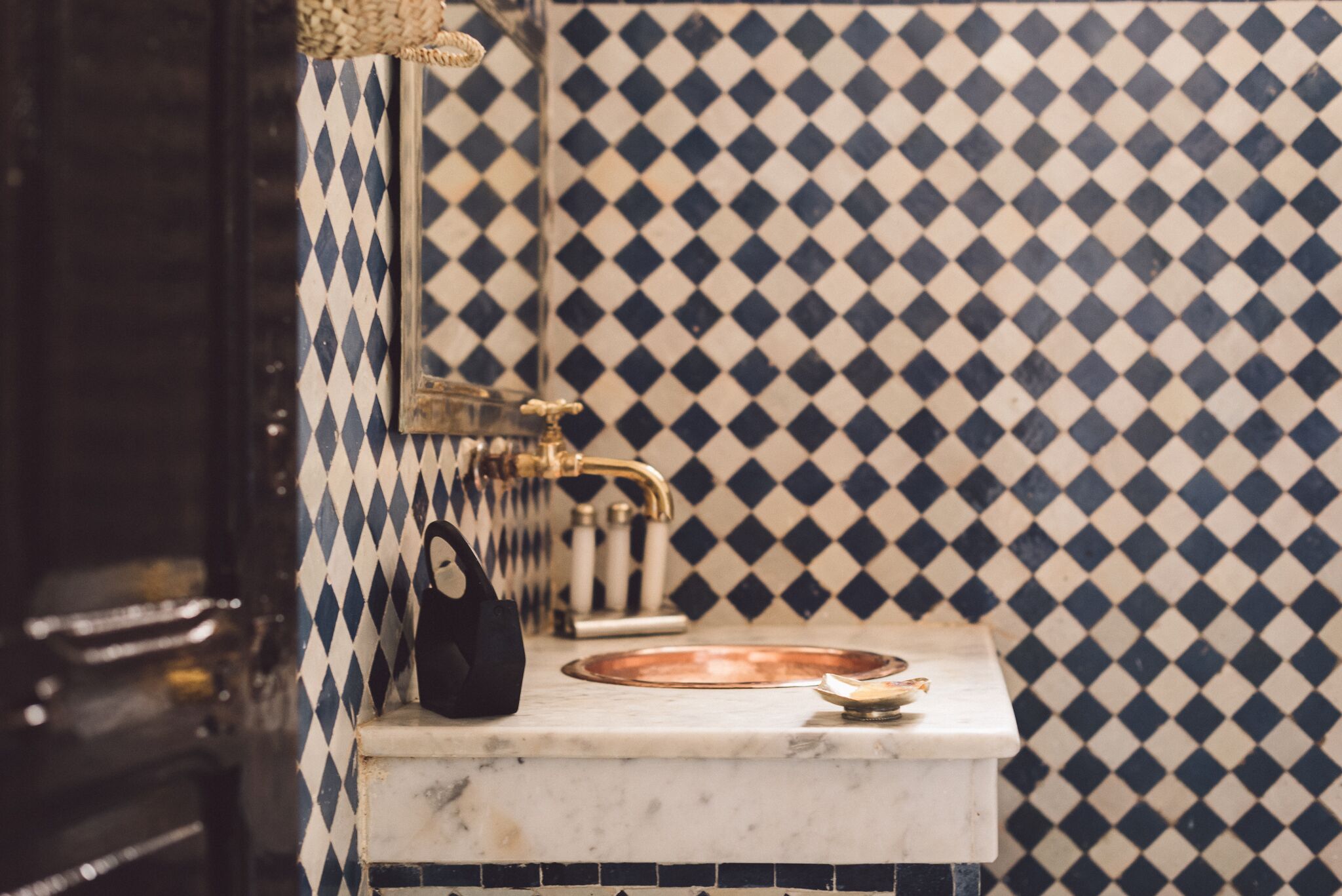 Um banheiro com revestimento de azulejos quadriculados, uma pia de cobre e uma bolsa preta no balcão.