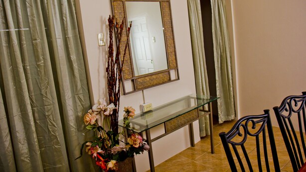Quarto com mesa de console de vidro, espelho e um vaso de flores. Há cortinas nas janelas e uma cadeira no canto.