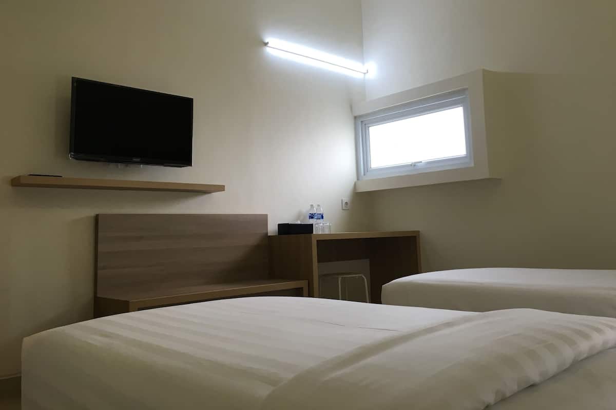 Quarto de hotel com duas camas, uma TV presa na parede, uma pequena escrivaninha, uma janela e uma luminária no teto.