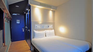 Quarto de hotel com cama, chão de madeira, porta azul e parede branca com luminária.
