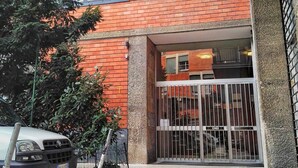 Entrada de um edifício com uma entrada por portão, um carro estacionado e uma árvore em primeiro plano.
