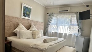 Uma cama bem arrumada com uma toalha enrolada, cabeceira com almofadas decorativas, uma obra de arte emoldurada acima da cama, uma televisão montada na parede e uma janela com cortinas.