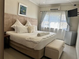 Uma cama bem arrumada com uma toalha enrolada, cabeceira com almofadas decorativas, uma obra de arte emoldurada acima da cama, uma televisão montada na parede e uma janela com cortinas.