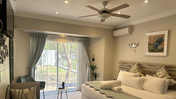 Quarto com cama, ventilador de teto, ar condicionado, janela com cortinas e uma pequena mesa com uma planta.