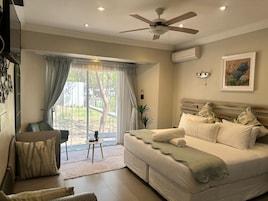Quarto com cama, ventilador de teto, ar condicionado, janela com cortinas e uma pequena mesa com uma planta.