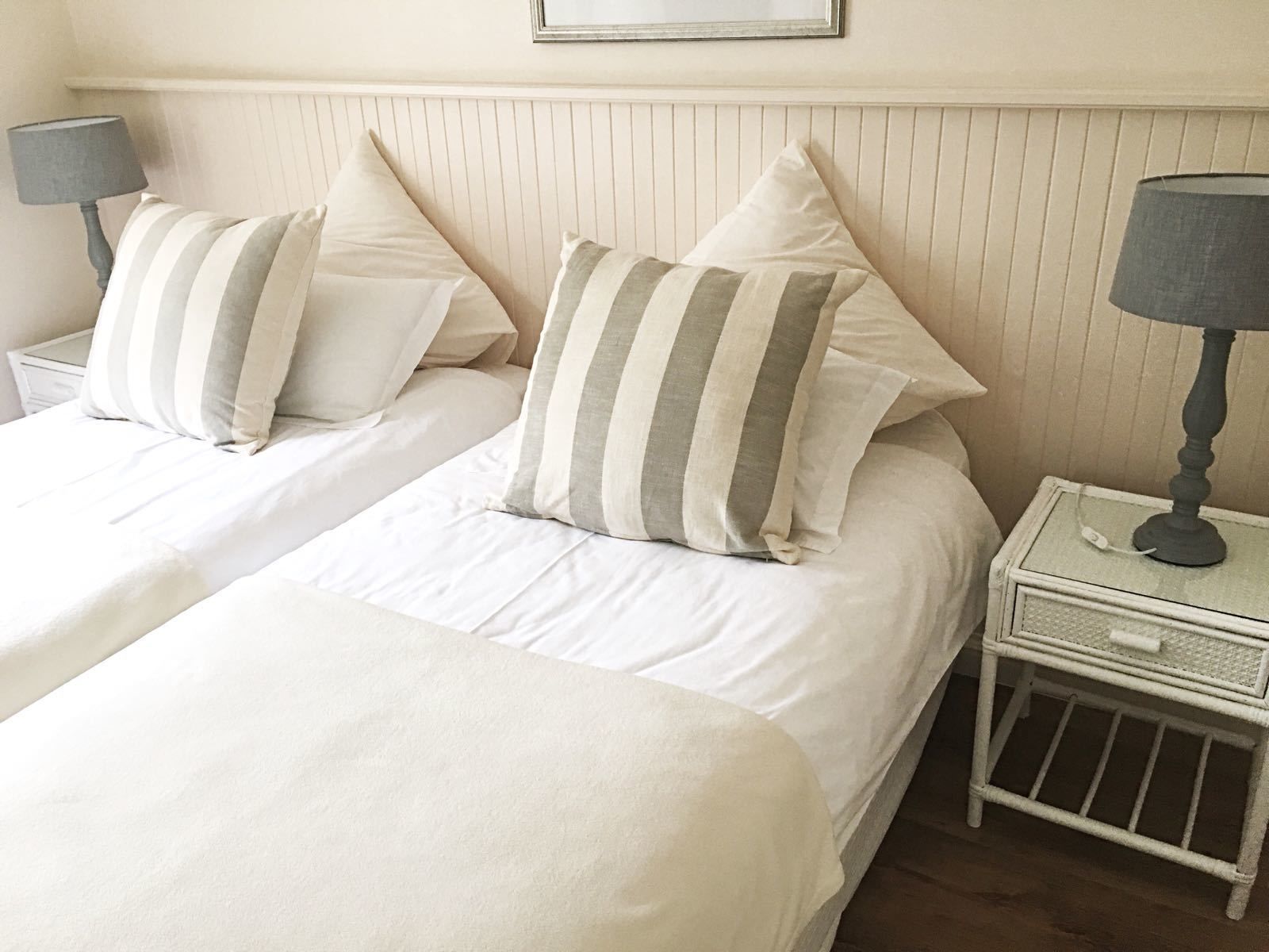 Quarto de hotel com duas camas, cada uma com roupa de cama branca e almofadas listradas. Há duas mesas de cabeceira com abajures.