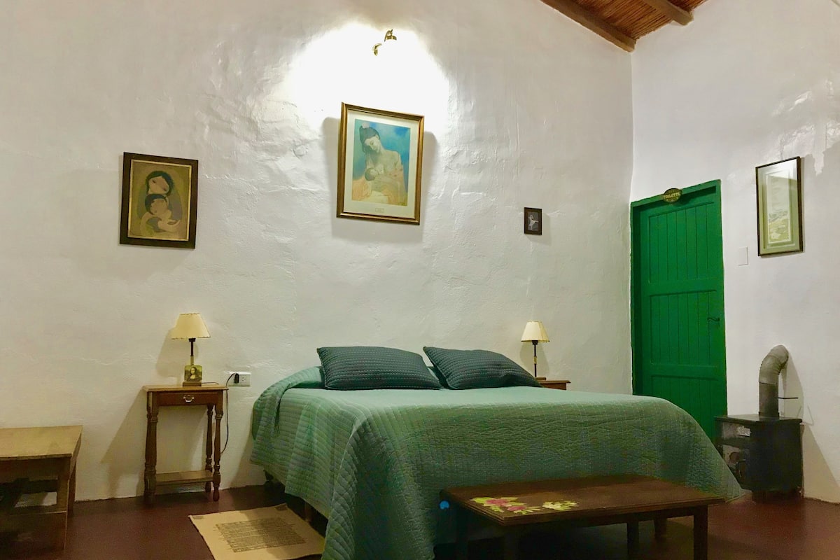 Quarto com uma cama, duas mesas de cabeceira, um banco, uma porta verde e uma parede com três quadros.