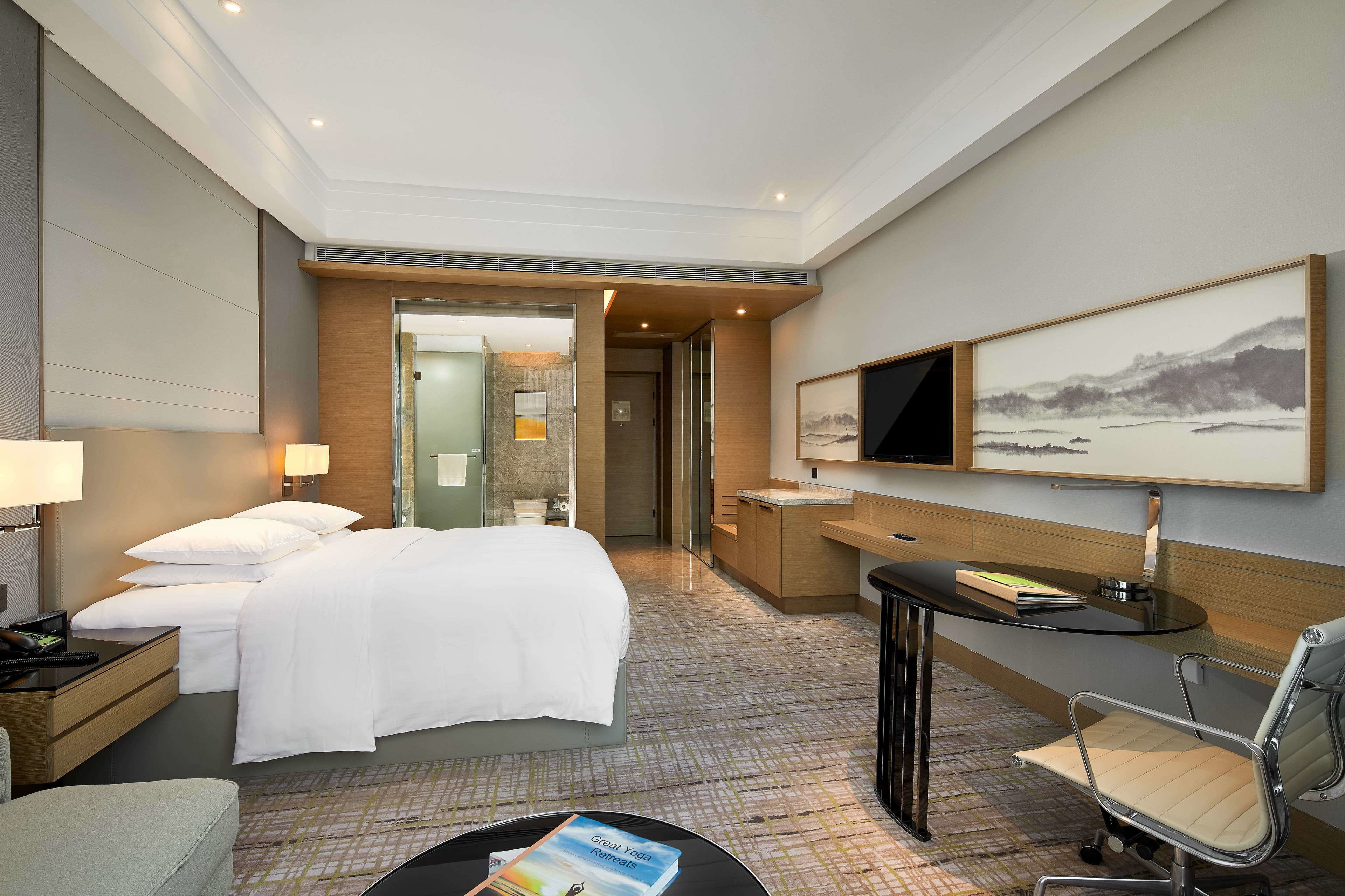 Quarto de hotel com cama, escrivaninha, cadeira, televisão e um quadro na parede.