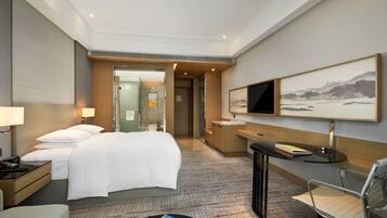 Quarto de hotel com cama, escrivaninha, cadeira, televisão e um quadro na parede.