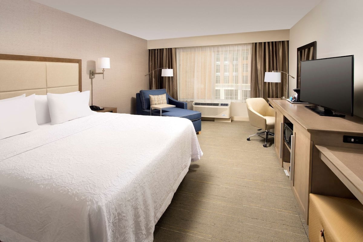 Quarto de hotel com uma cama grande, uma escrivaninha com televisão de tela plana, uma cadeira e uma luminária de mesa.