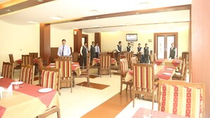 Uma área de refeições de hotel com várias mesas e cadeiras, uma tela de televisão e um grupo de pessoas ao fundo.