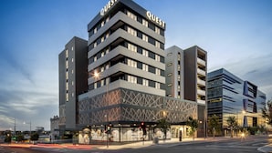 Um hotel de vários andares com a sinalização 'QUEST', fachada moderna e vista da rua ao entardecer.