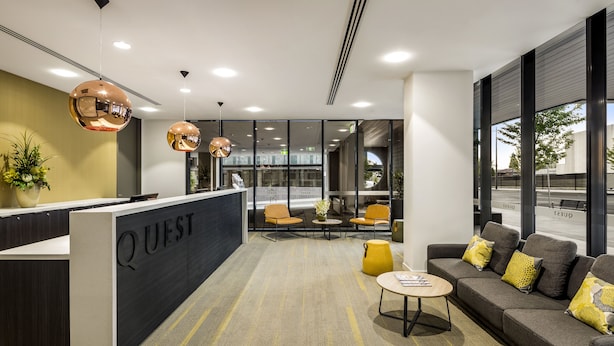 Uma área de recepção moderna com uma placa 'QUEST', assentos e vista através de grandes janelas.