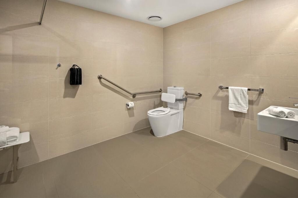 Um banheiro moderno com azulejos bege, vaso sanitário branco, pia com porta-toalhas e corrimão para acessibilidade.
