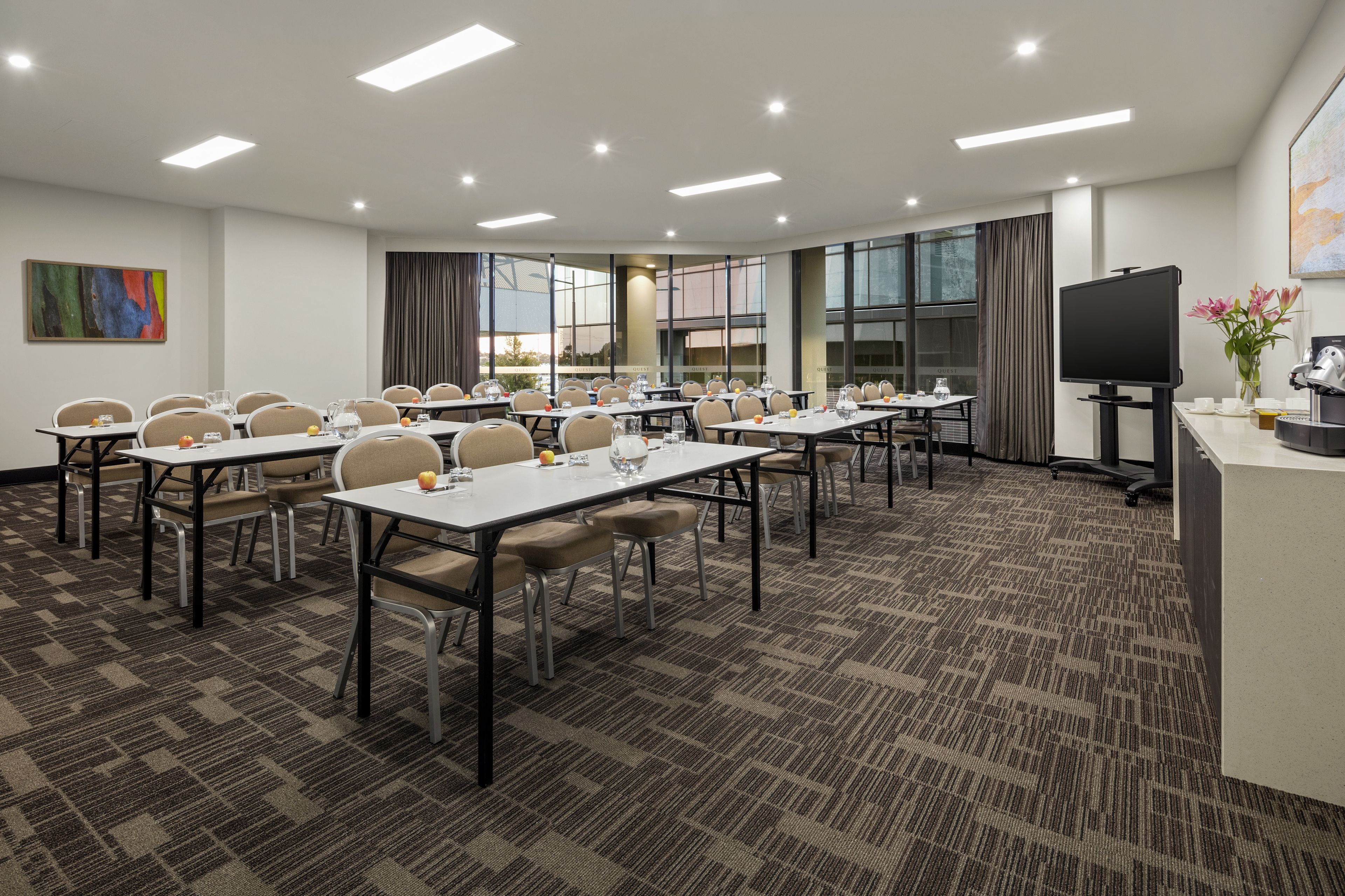 Sala de conferências com várias mesas e cadeiras, uma televisão e uma janela grande com cortinas.
