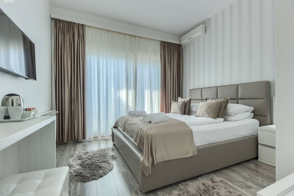 Quarto moderno com uma cama grande, mesa de cabeceira, televisão de tela plana e uma janela ampla com cortinas.