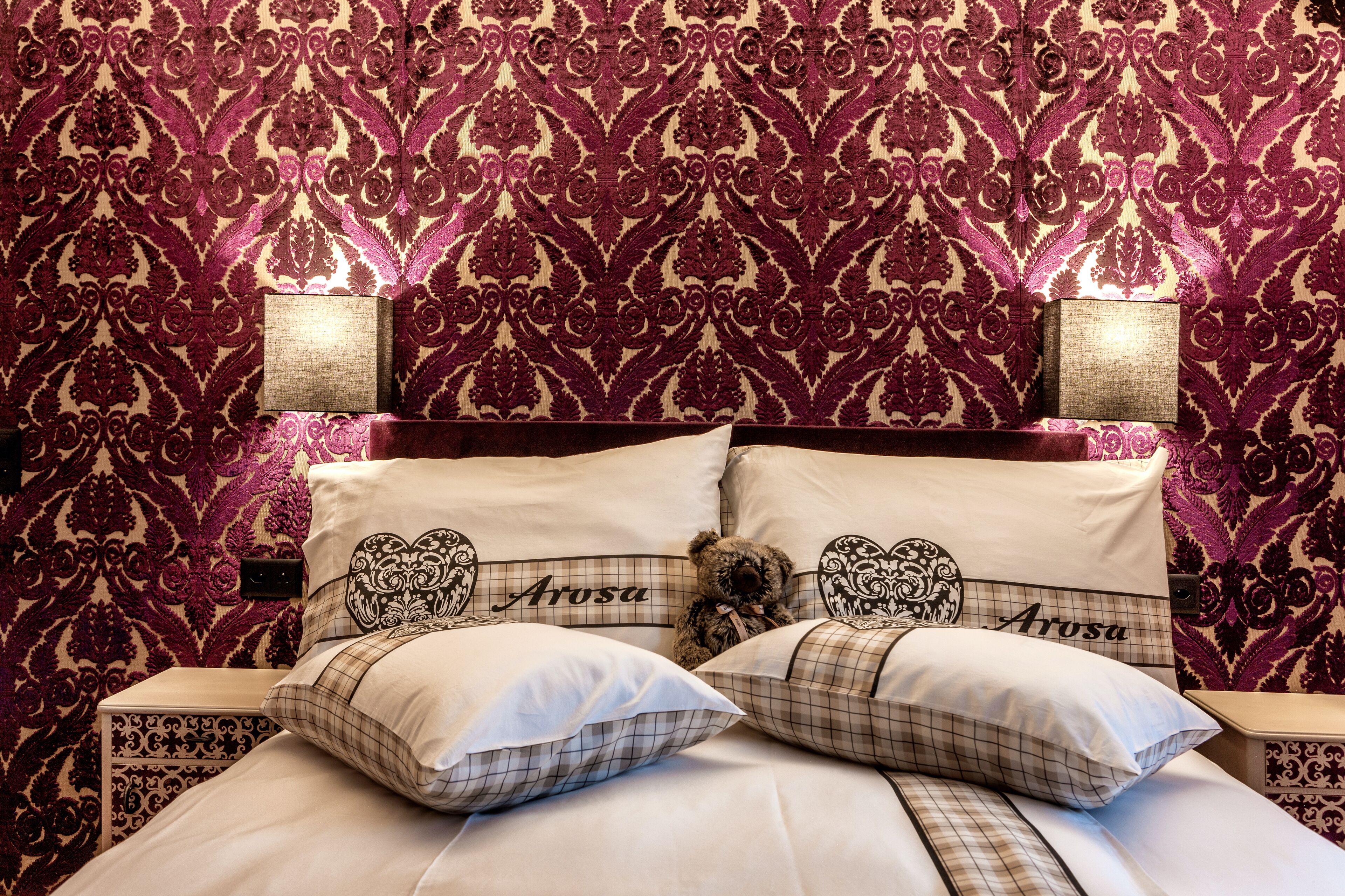 Uma cama com duas almofadas, uma delas com um desenho de coração e a palavra "Arosa", e um ursinho de pelúcia sobre a cama.