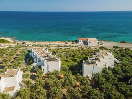 Vista aérea de uma área residencial costeira com edifícios brancos, uma estrutura pequena com telhado vermelho e uma vista clara do mar.