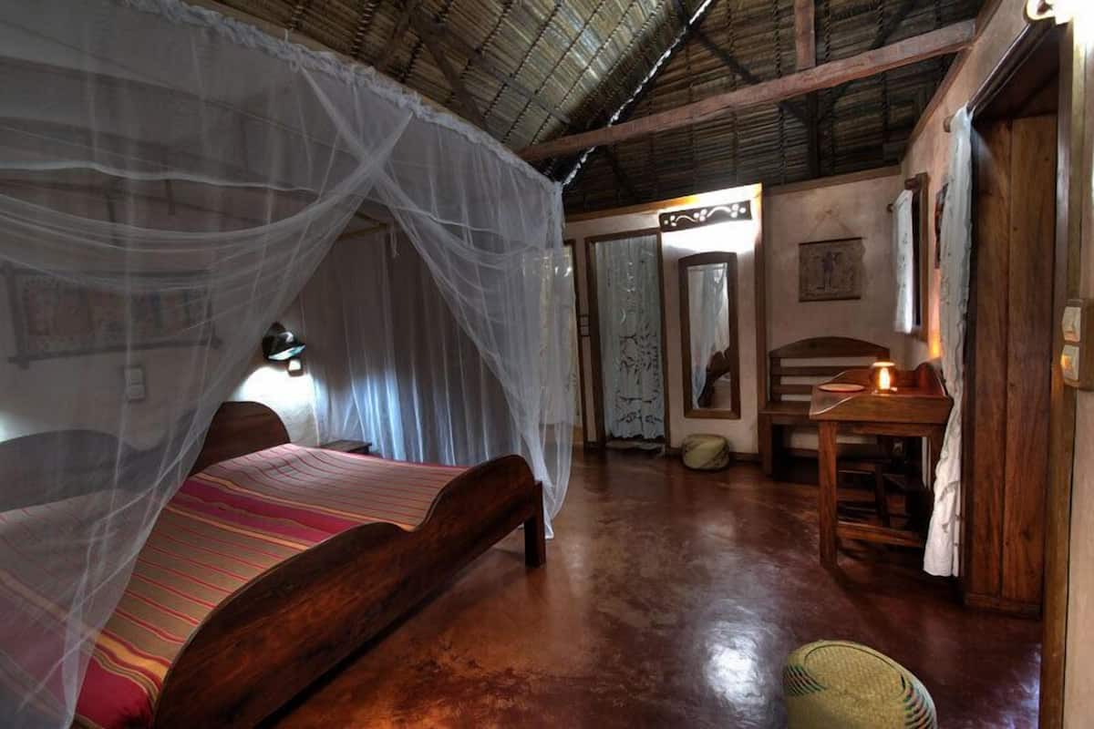 Quarto com cama sob um mosquiteiro, uma escrivaninha de madeira com luminária e um espelho na parede.