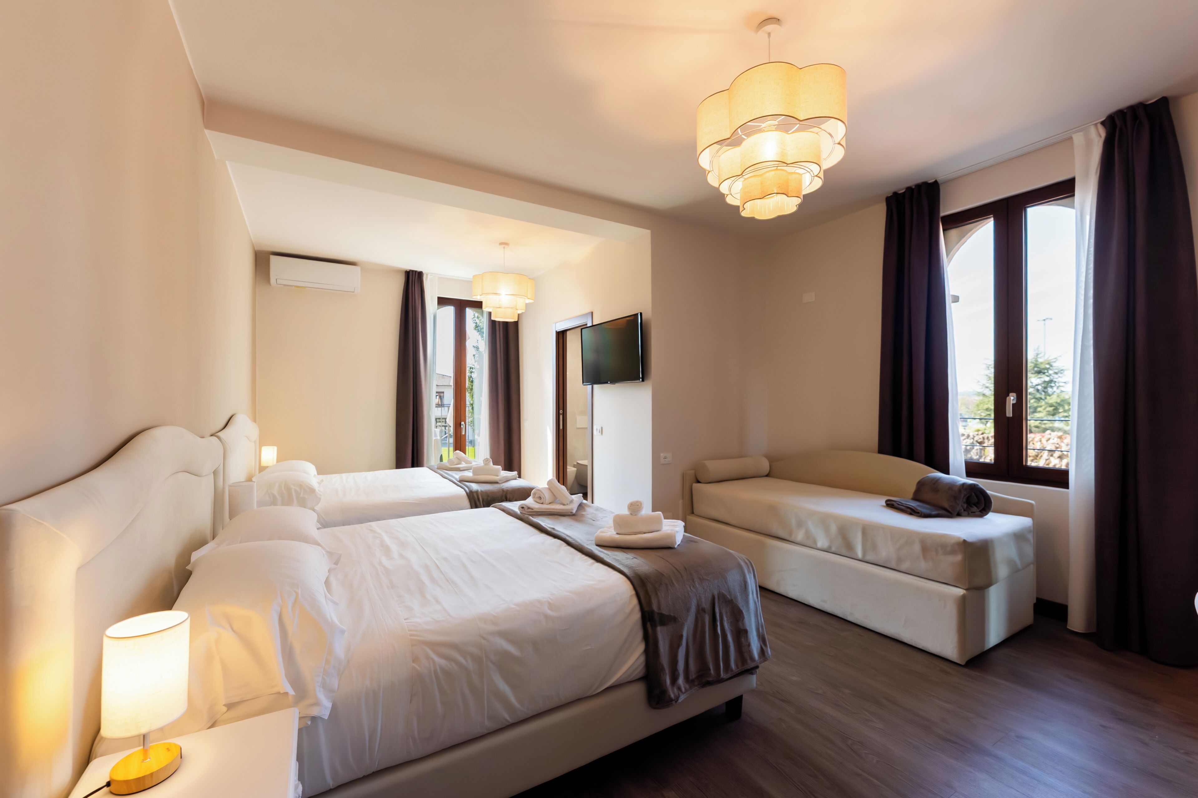 Quarto de hotel com duas camas, um sofá, uma televisão e uma janela grande com cortinas.