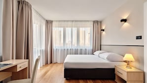 Quarto de hotel com cama, mesa de cabeceira com abajur, escrivaninha com cadeira e cortinas.