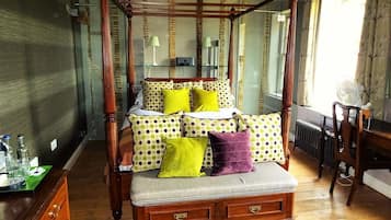 Uma cama com dossel sobre pilares, com estrutura de madeira, um banco com almofadas e uma pequena mesa com garrafas.