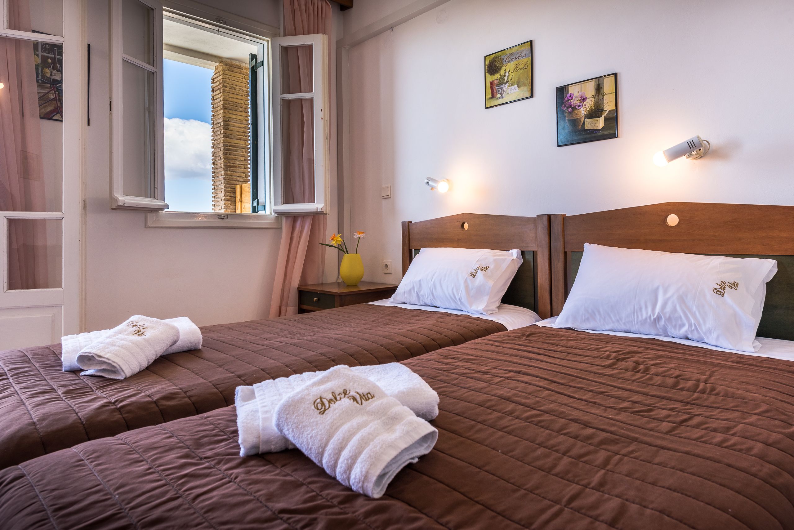 Quarto de hotel com duas camas, cada uma com colcha acolchoada marrom e almofadas brancas. Há uma janela com vista para o céu e nuvens, e um candeeiro fixado na parede.