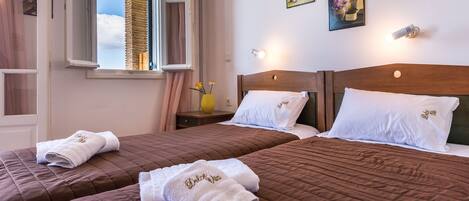 Quarto de hotel com duas camas, cada uma com colcha acolchoada marrom e almofadas brancas. Há uma janela com vista para o céu e nuvens, e um candeeiro fixado na parede.