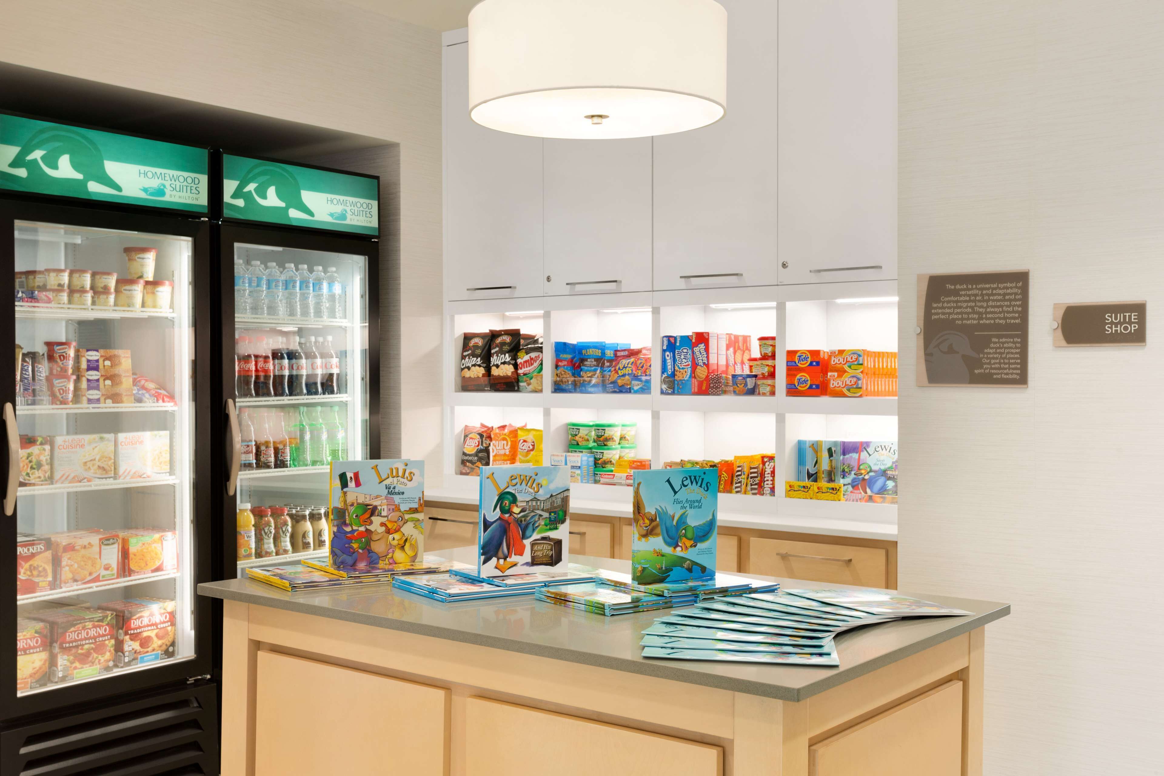 Uma área de recepção de hotel com um balcão exibindo livros e revistas, uma geladeira repleta de bebidas e lanches, e uma placa indicando a localização da loja de suítes.