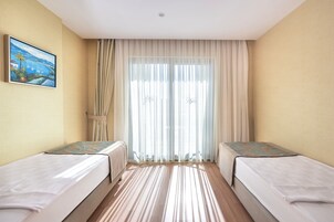 Quarto de hotel com duas camas de solteiro, uma janela grande com cortinas leves, um quadro na parede e piso de madeira.