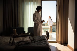 Quarto de hotel com uma cama, um serviço de mesa com itens de café da manhã e vista para uma varanda com uma pessoa lá fora.