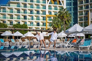 Uma área de piscina de hotel com espreguiçadeiras, guarda-sóis e o reflexo do edifício na água.