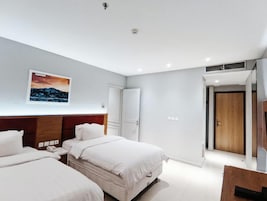 Quarto de hotel com duas camas, cabeceira de madeira, um quadro na parede, uma porta e uma escrivaninha.