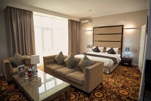 Quarto de hotel com sofá, mesa de centro de vidro, cama com cabeceira, mesa de cabeceira e tapete estampado.