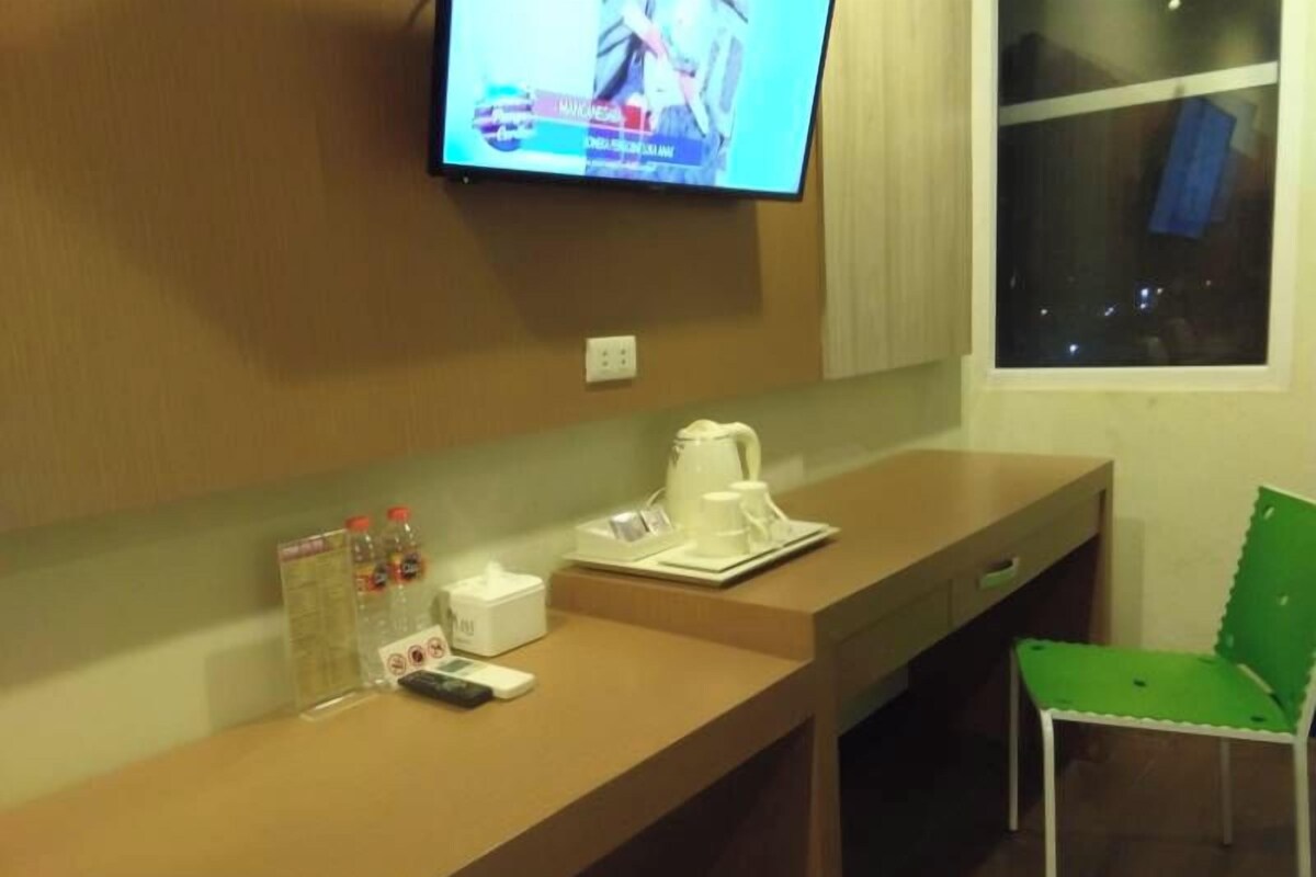 Quarto de hotel com uma escrivaninha de madeira, televisão presa à parede, janela com vista noturna, uma cadeira verde e uma cafeteira.