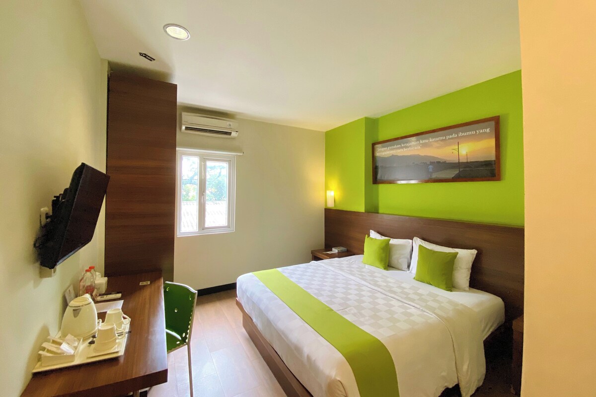 Quarto de hotel com cama, escrivaninha, cadeira, televisão e janela.