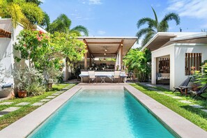 Piscina com um terraço coberto, rodeada por vegetação exuberante e palmeiras.