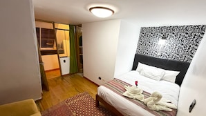 Quarto de hotel com uma cama, uma pequena mesa com uma vela, uma cadeira, um tapete estampado e uma parede com padrão de folhas.