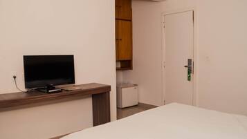 Quarto de hotel com uma cama, uma televisão sobre um móvel de madeira, um frigorífico pequeno e uma porta com um sinal verde.
