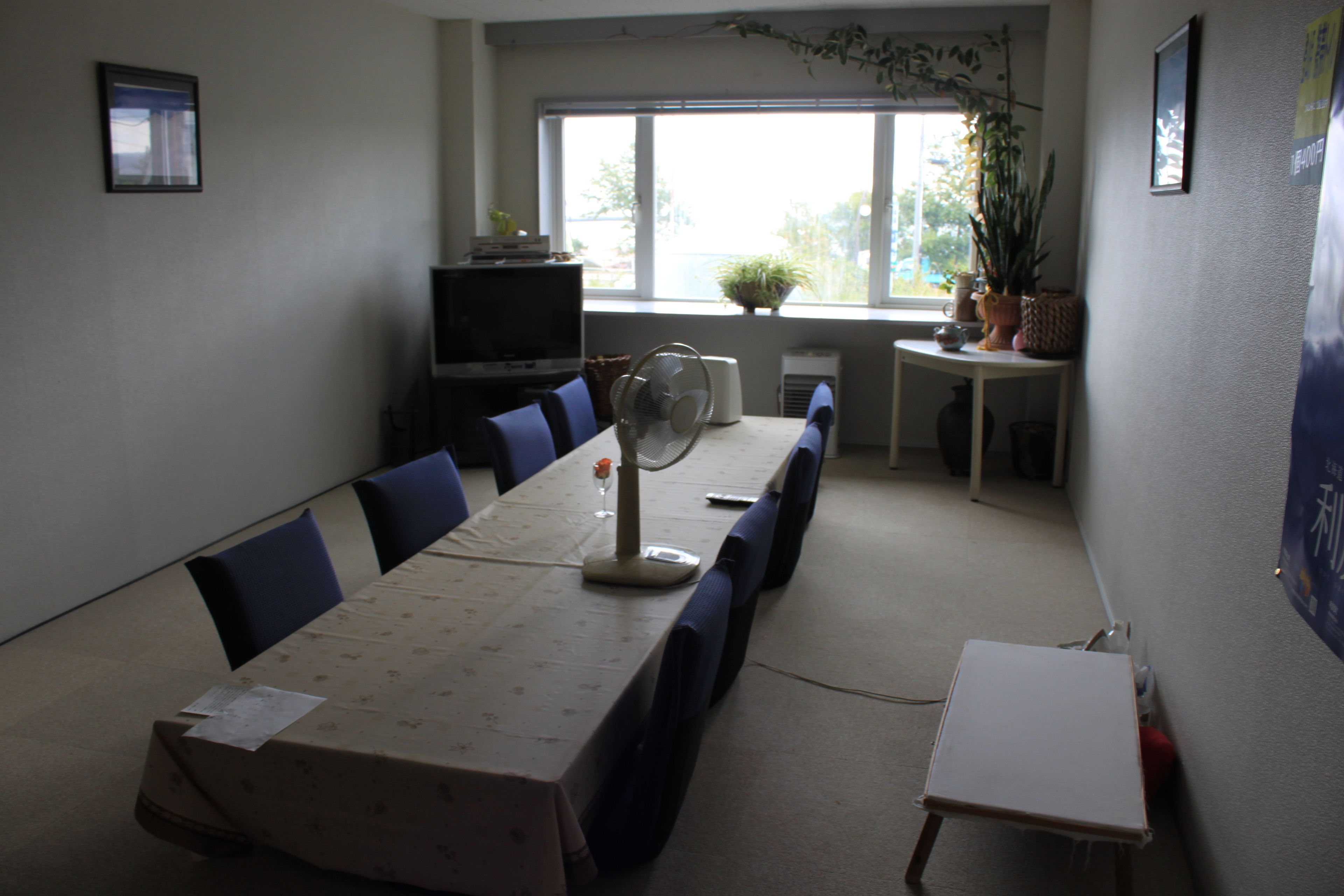 Uma sala de conferências com uma mesa comprida, cadeiras, um ventilador, uma TV e uma janela com plantas.