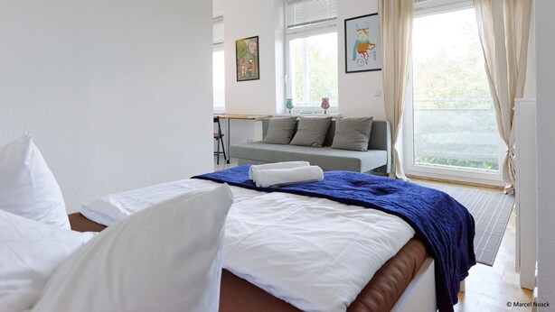Quarto de hotel com cama, sofá, escrivaninha e janela com cortinas.