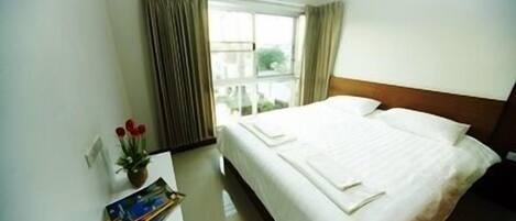Quarto de hotel com uma cama, uma mesinha de cabeceira com um livro e um vaso de flores, e uma janela com cortinas.