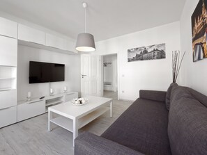 Sala de estar moderna com sofá branco, mesa de centro branca, televisão de tela plana e luminária pendente. Há uma porta que dá para outro cômodo e um quadro na parede.