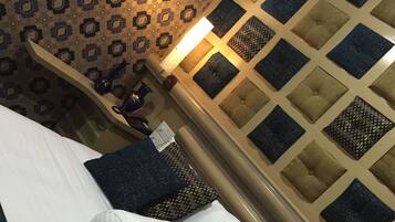 Quarto de hotel com teto estampado, cama com roupa de cama branca e parede com painéis decorativos quadrados.