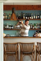 Uma área de bar com prateleiras de madeira repletas de garrafas de bebidas, uma luminária pendente e cadeiras de vime.