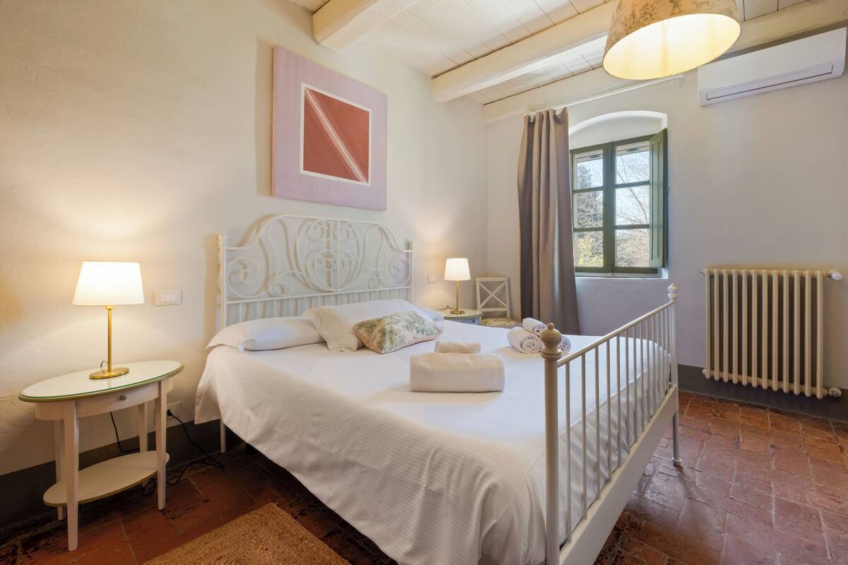 Quarto com cama, luminárias de cabeceira, radiador e janela com cortinas.